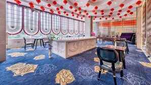 Um saguão de hotel com balcão de recepção, área de estar e grandes janelas com cortinas decorativas.