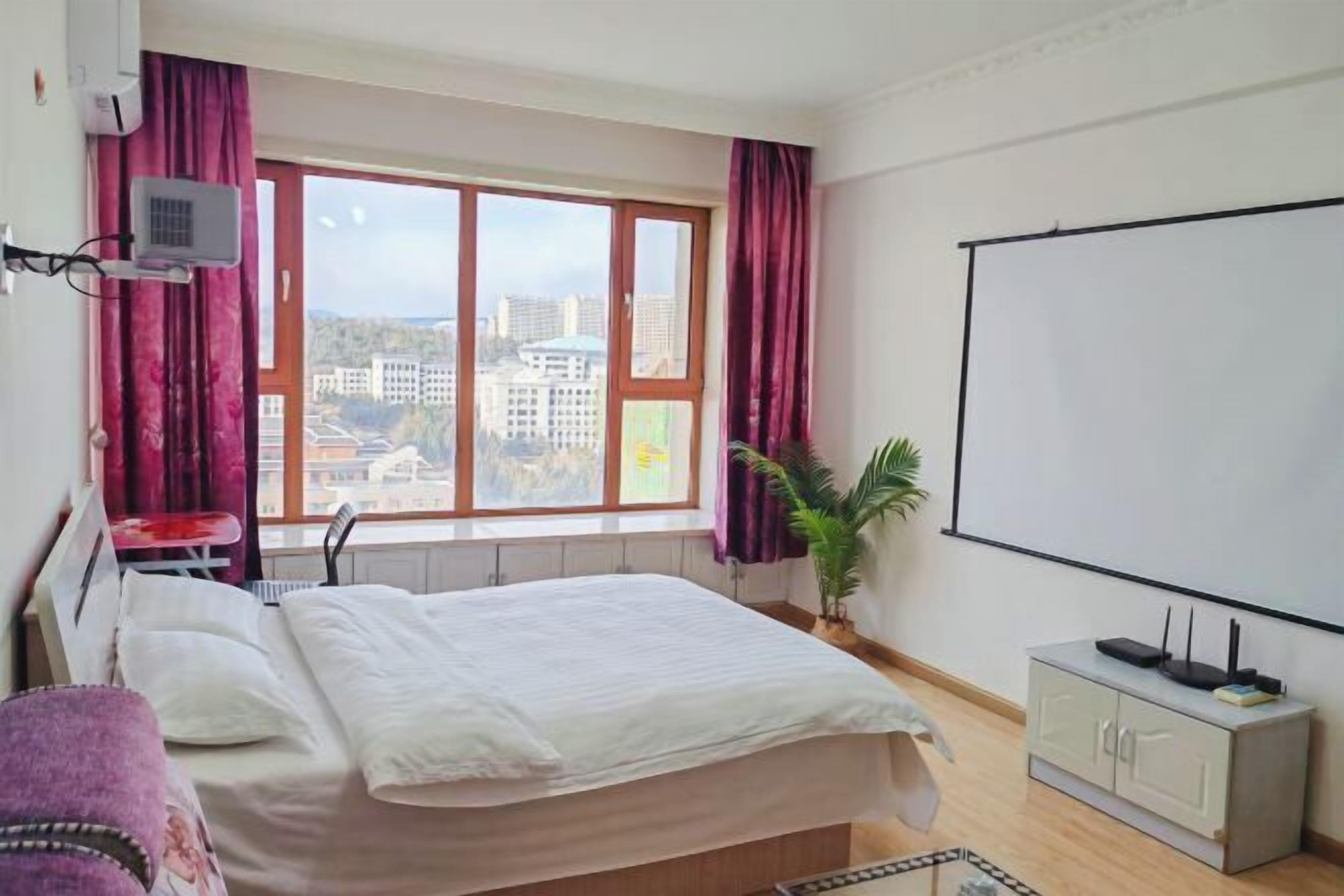 Quarto de hotel com cama, escrivaninha, janela com cortinas, planta e tela de projetor.