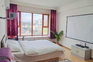 Quarto de hotel com cama, escrivaninha, janela com cortinas, planta e tela de projetor.