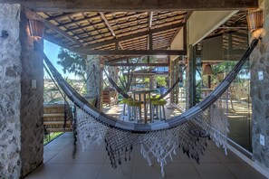 Uma rede sob uma pérgola de madeira, uma parede de pedra e uma porta de vidro que dá para um pátio ao ar livre com uma mesa e cadeiras.
