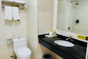 Casa de banho com sanita branca, lavatório e espelho. Há um porta-toalhas com toalhas, um contentor de lixo e uma pequena prateleira com artigos de higiene pessoal.
