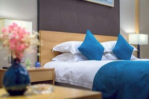 Quarto de hotel com cabeceira de madeira, roupa de cama branca e almofadas azuis. Há um vaso com flores na mesinha de cabeceira e uma luminária na mesa lateral.
