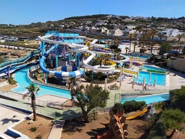 Um parque aquático com vários toboáguas e piscinas, cercado por palmeiras e uma paisagem montanhosa ao fundo.