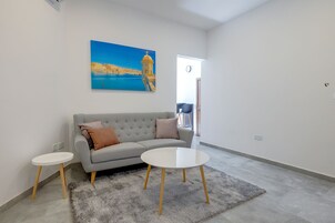 Uma sala de estar moderna com um sofá cinza, duas mesinhas de cabeceira brancas e um tapete felpudo cinza.