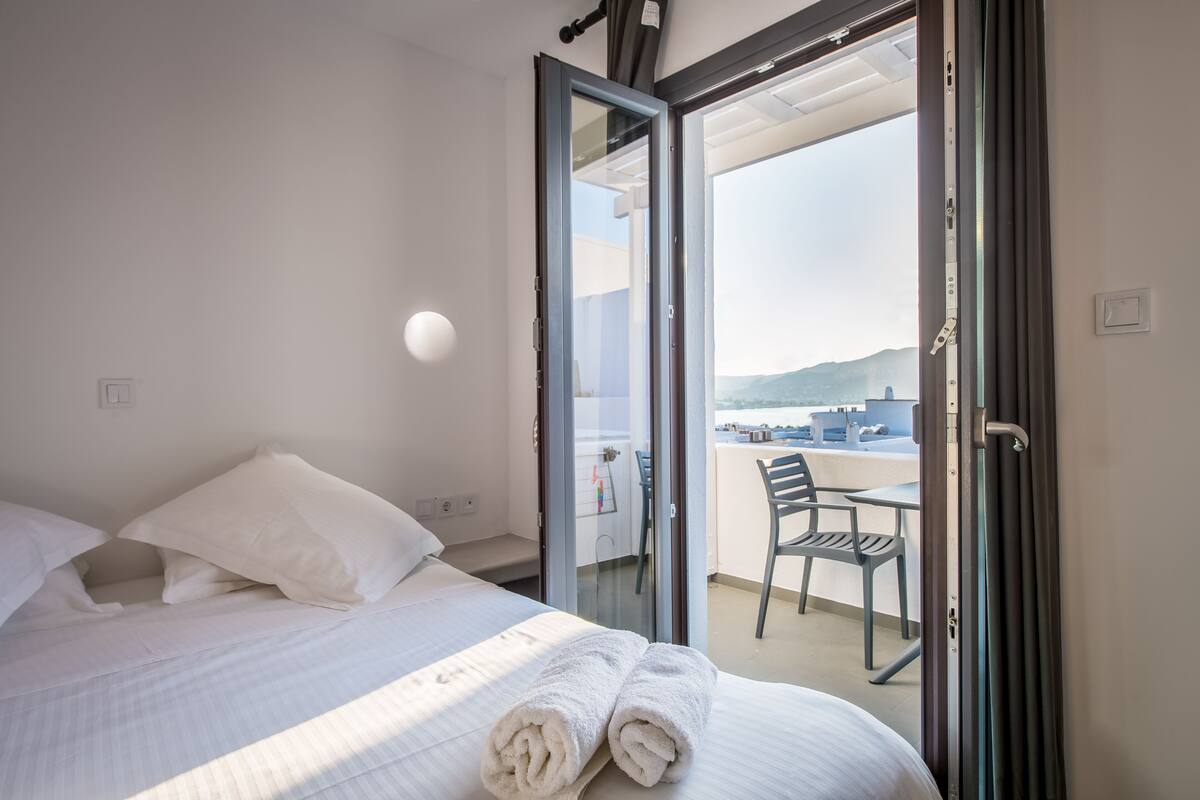 Quarto de hotel com cama, varanda com mesa e cadeiras, e vista para o mar.