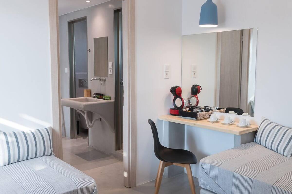 Quarto de hotel moderno com cama, escrivaninha com cadeira, espelho e pequena cozinha.