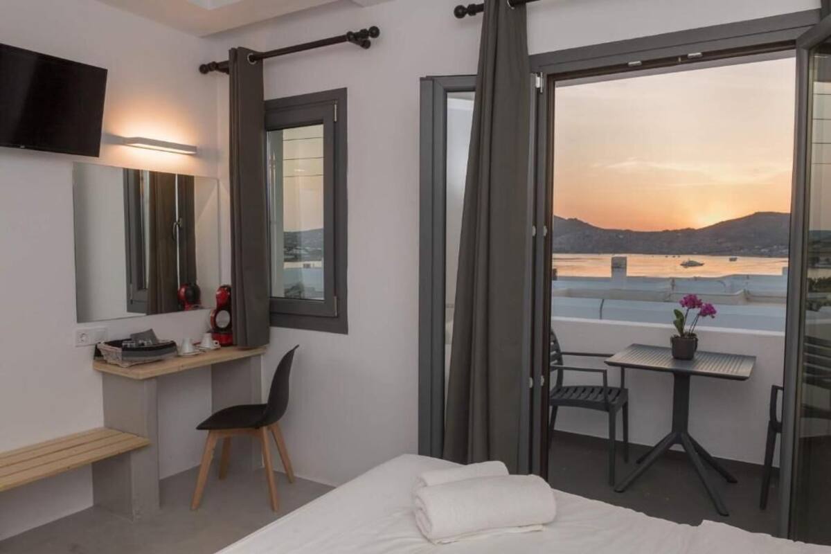 Quarto de hotel moderno com cama, escrivaninha, poltrona, televisão e varanda com vista para o mar e montanhas.