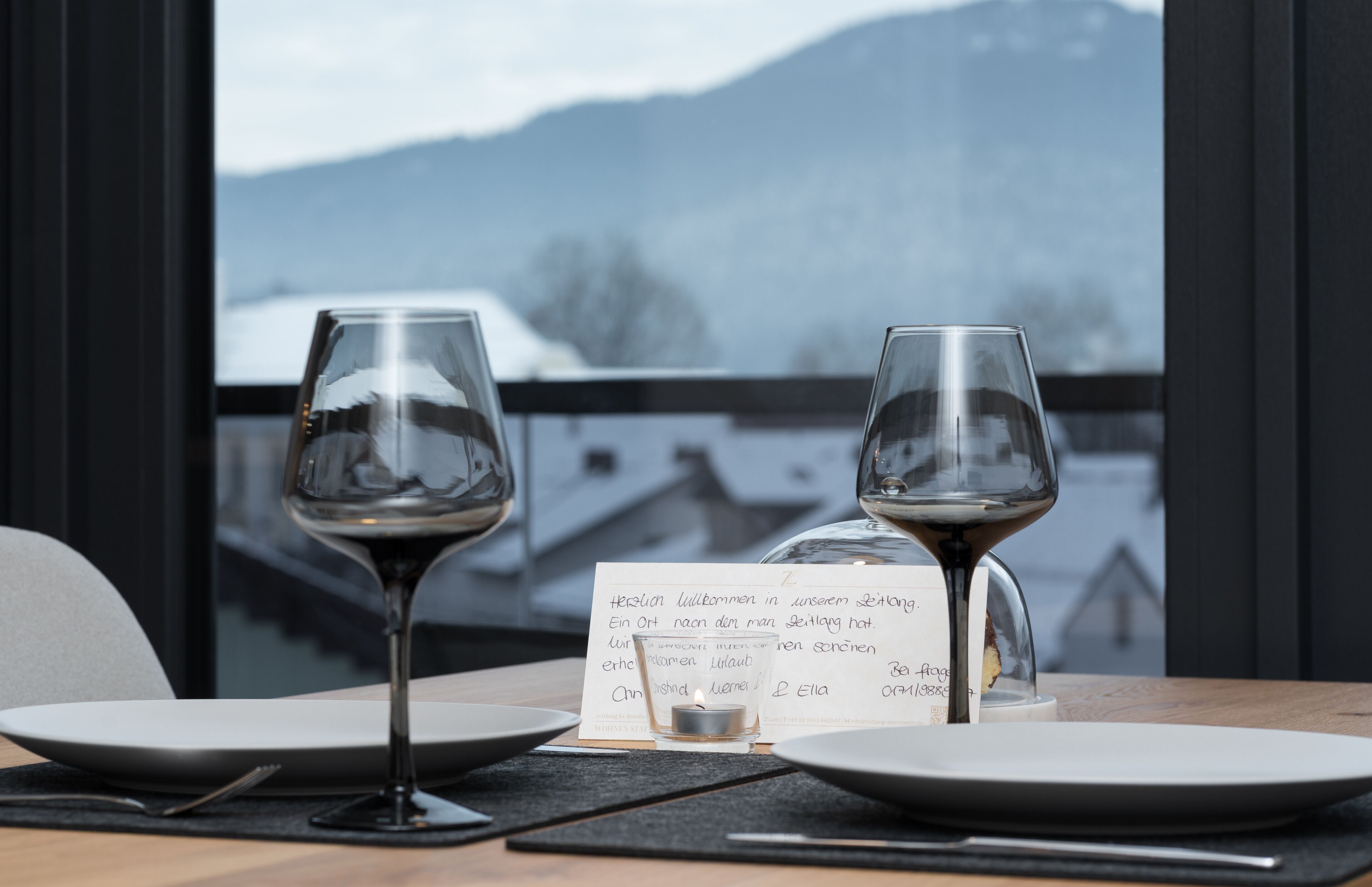 Mesa posta para dois, com taças de vinho e um cardápio, com vista para uma paisagem montanhosa coberta de neve.