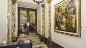 Um amplo salão com colunas de mármore, uma escadaria e um grande quadro emoldurado na parede.