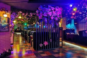 Uma área de bar com piso xadrez, divisória preta com flores e decorações florais penduradas.