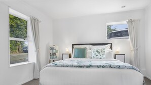 Quarto com uma cama grande, duas mesas de cabeceira, uma janela com cortinas e uma placa com palavras motivacionais.