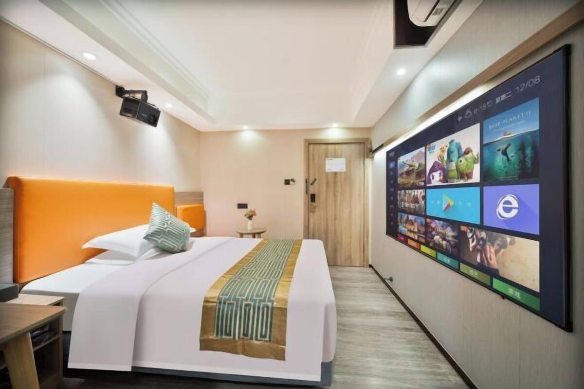 Quarto de hotel com cama, escrivaninha, parede com televisão e porta.