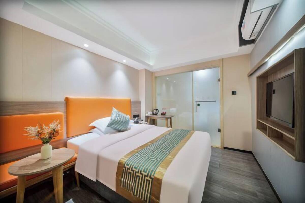 Quarto de hotel com cama, cabeceira laranja, mesa de cabeceira com um vaso, televisão e guarda-roupa.