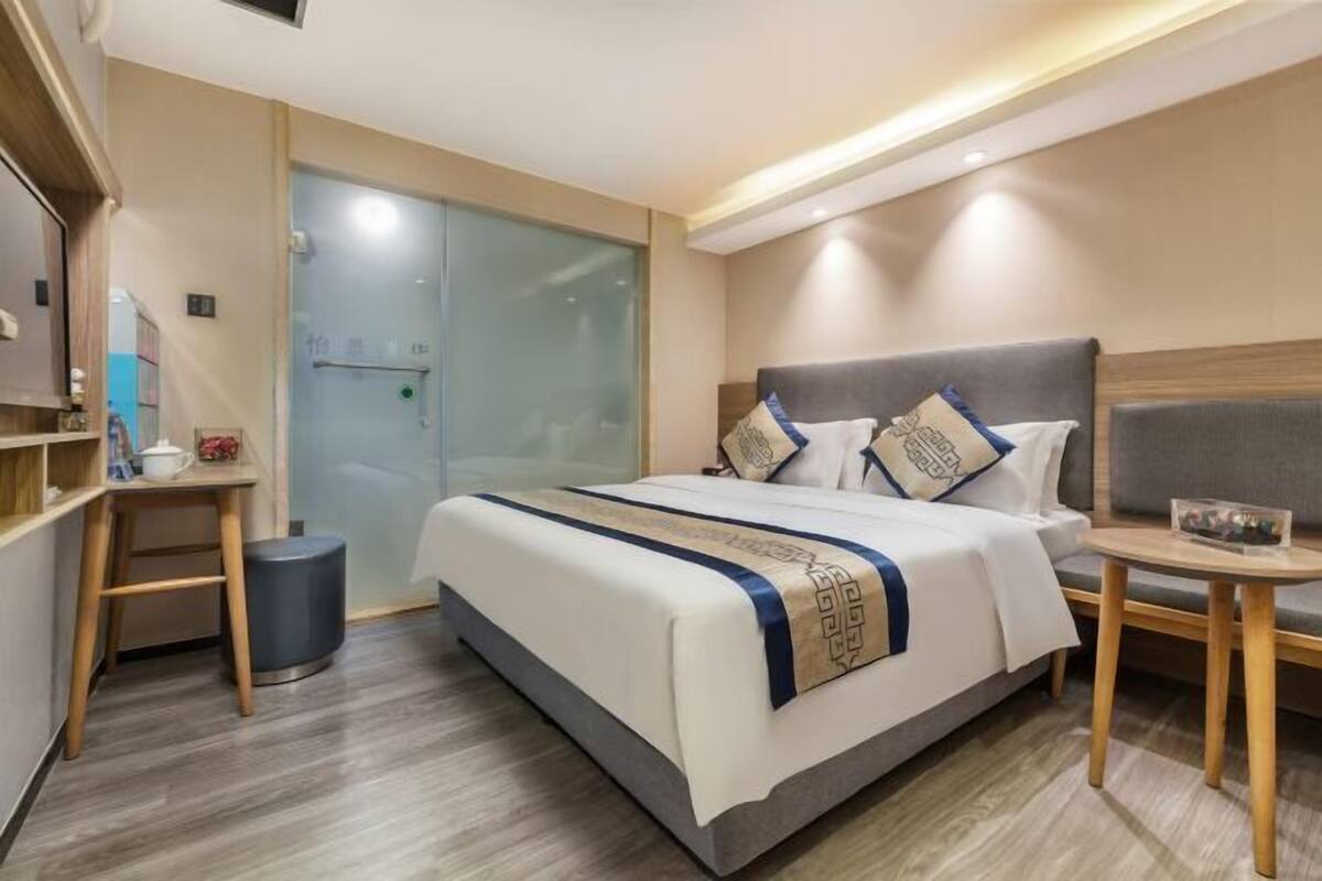 Quarto de hotel com cama, mesas de cabeceira, um banquinho, lixeira e porta de vidro que dá para um banheiro.