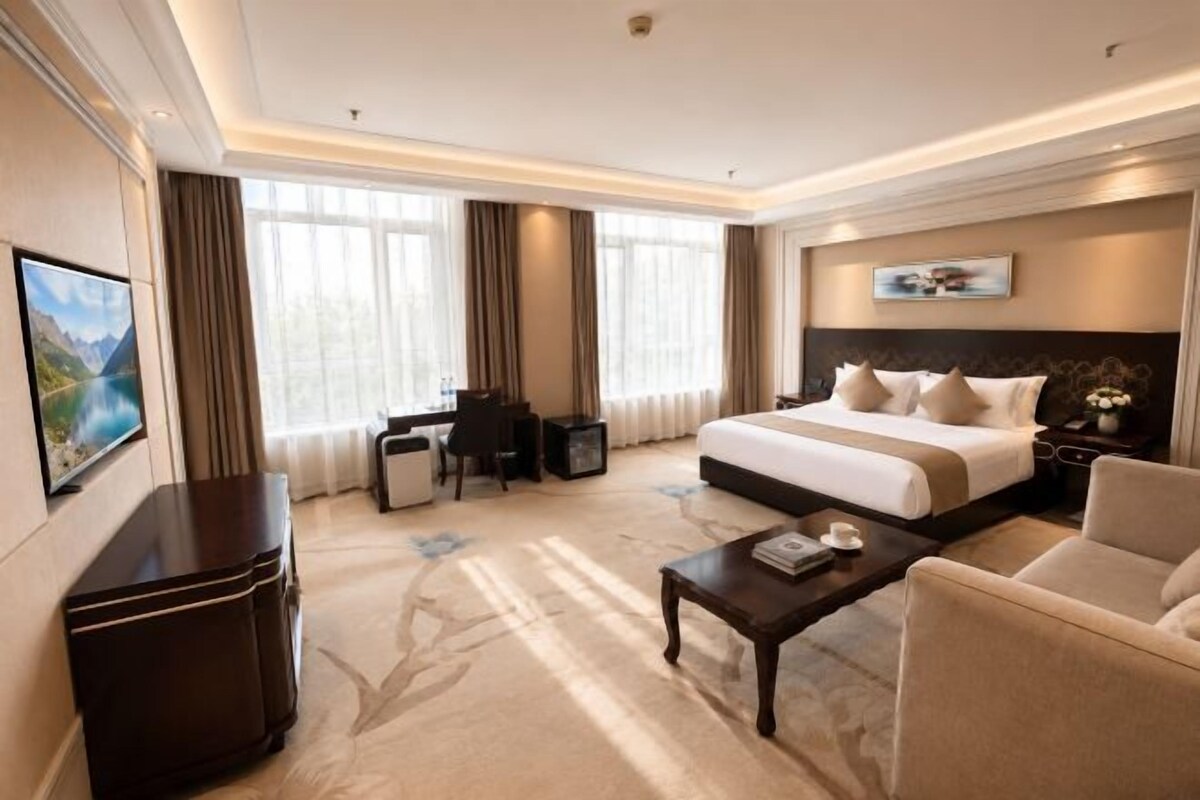 Quarto de hotel com cama grande, escrivaninha, cadeira, mesa de centro e televisão.