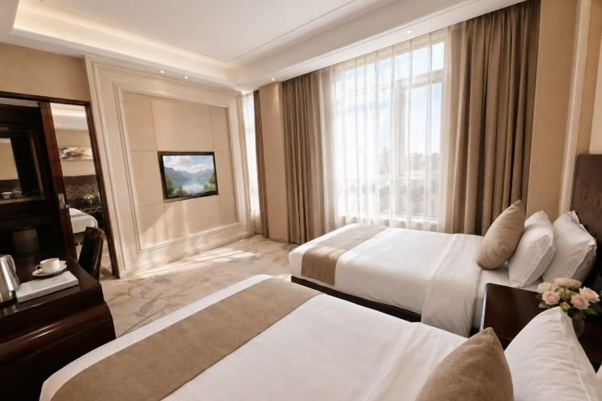 Quarto de hotel com duas camas, televisão, escrivaninha e uma janela grande com cortinas.