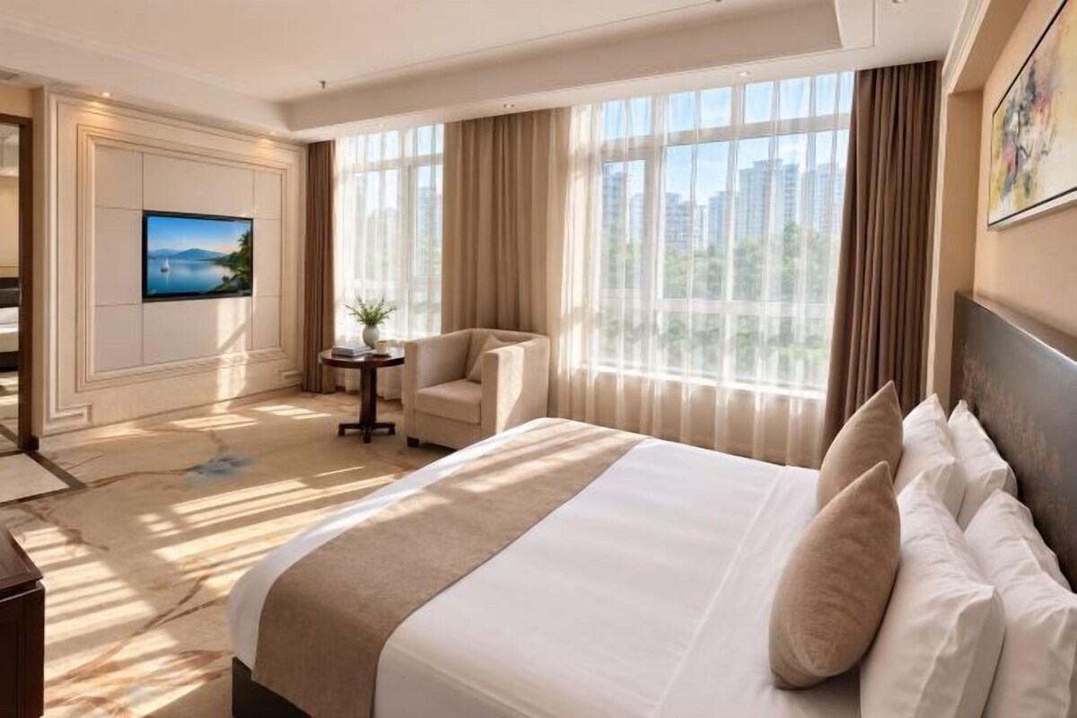 Quarto de hotel com uma cama grande, área de estar com uma cadeira e mesa, e vista da cidade por grandes janelas.