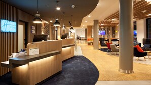 Uma área de recepção de hotel moderna, com um balcão de madeira, várias luminárias pendentes e um espaço de estar com cadeiras e mesas.