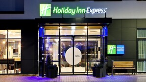 Exterior de um hotel Holiday Inn Express com uma grande entrada de vidro, um letreiro com a palavra 'Hello' e um banco na frente.