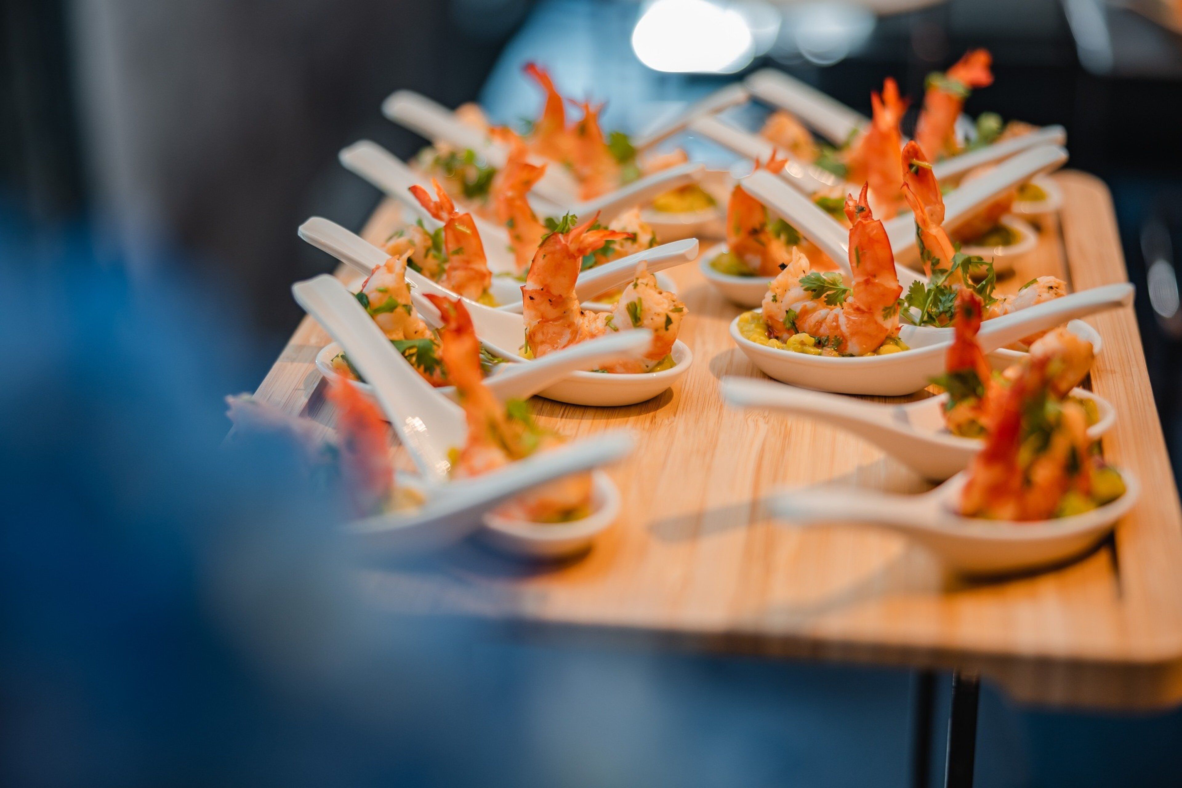 Uma mesa com colheres brancas servindo camarões como aperitivo.