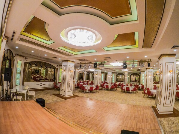 Uma sala de banquetes com mesas redondas, cadeiras vermelhas e colunas decoradas.