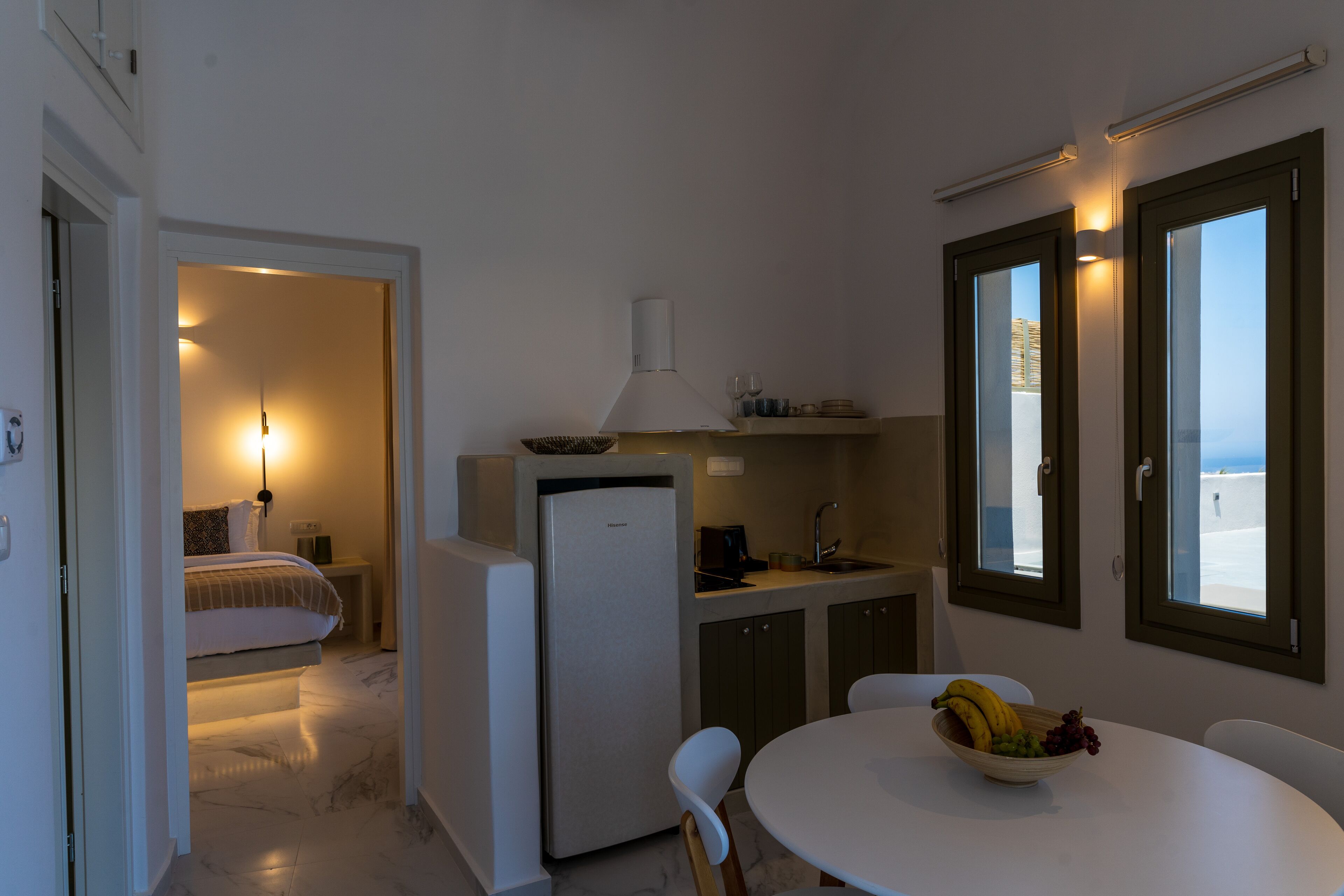 Quarto de hotel compacto com cama, pequeno espaço de cozinha, área de refeições com mesa e cadeiras, e vista pelas janelas.