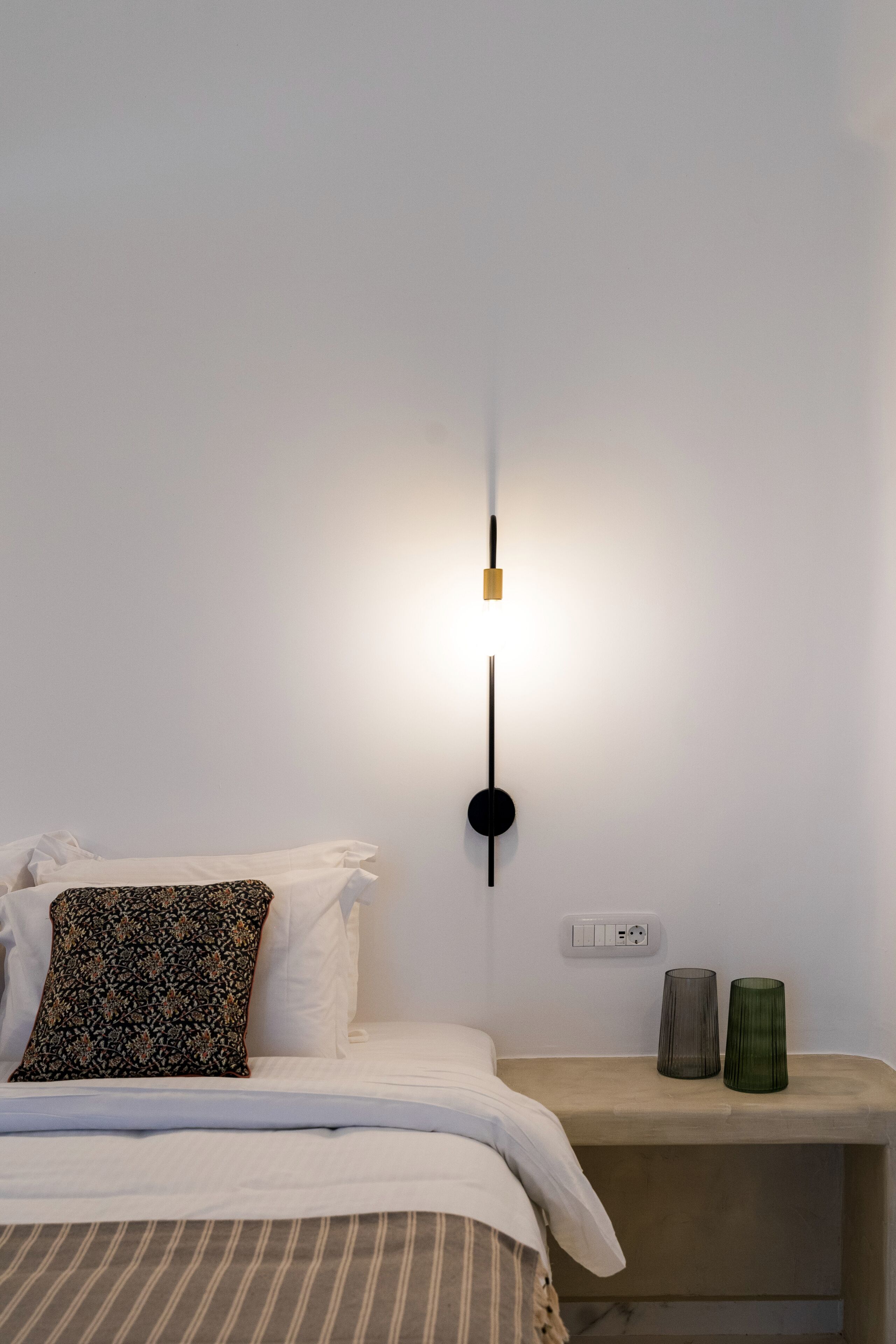 Cama bem arrumada com almofada estampada, mesa de cabeceira com dois vasos e luminária fixada na parede.