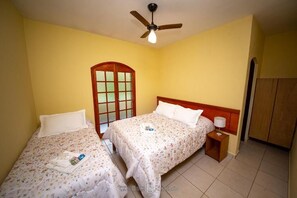 Quarto de hotel com duas camas, um guarda-roupa de madeira, um ventilador de teto e uma janela com moldura de madeira.