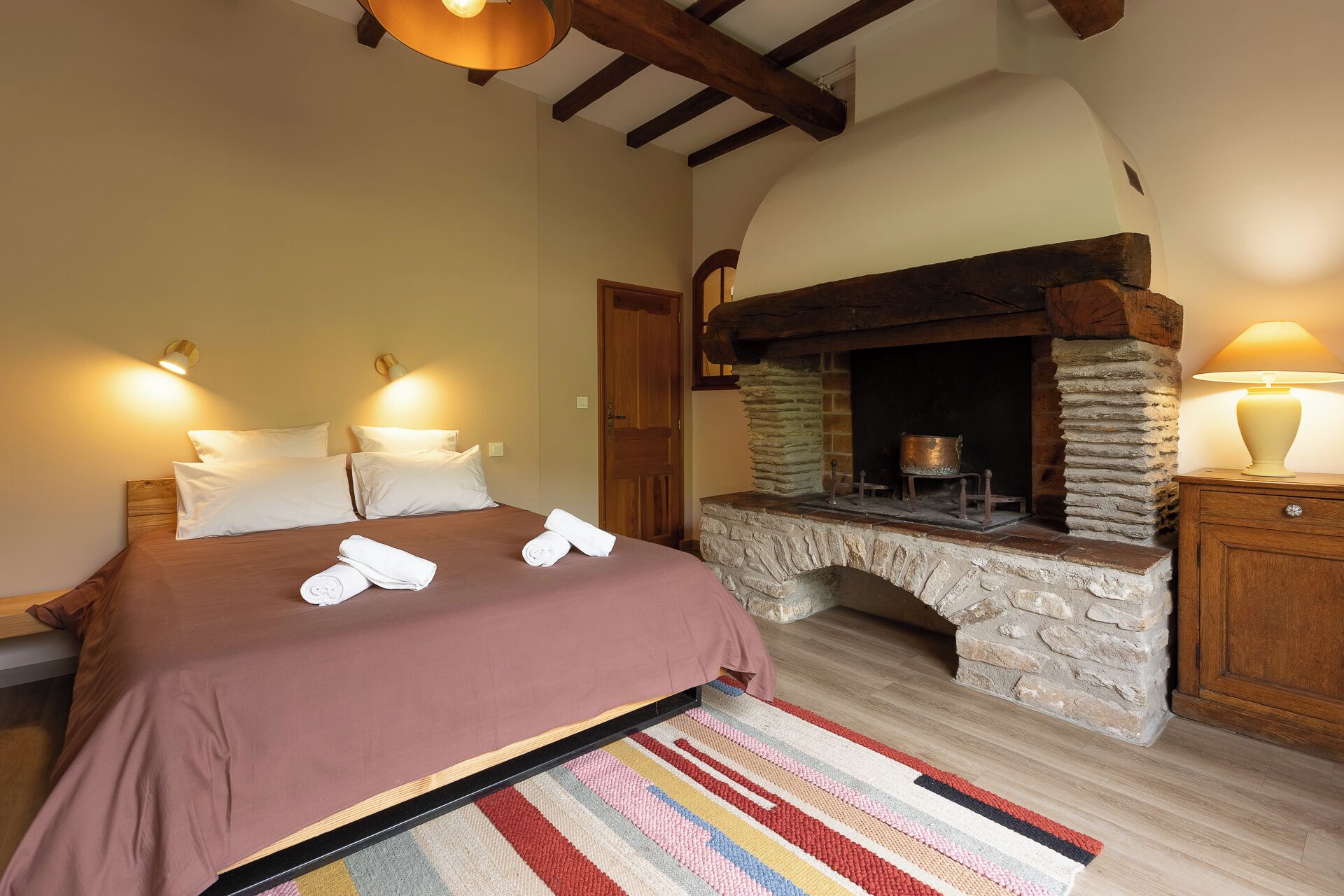 Quarto com lareira de pedra, cama com colcha marrom e tapete listrado colorido.