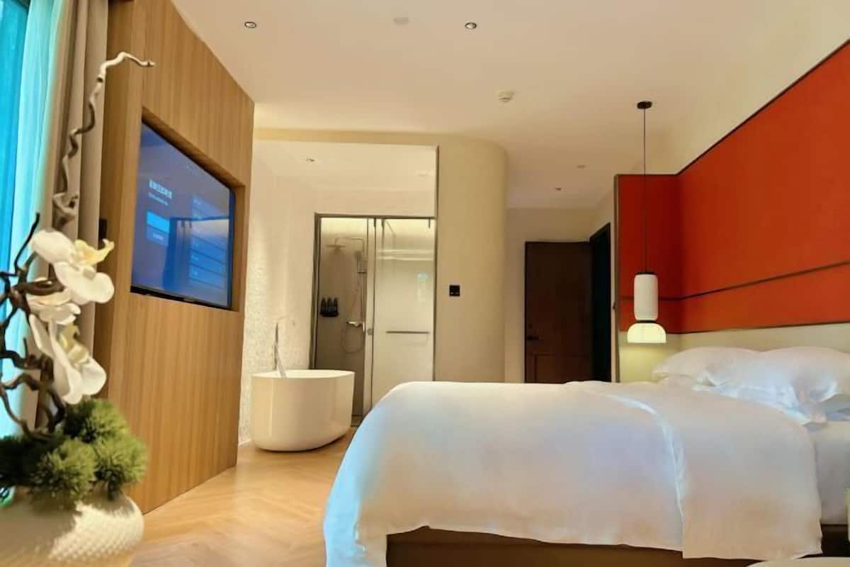Quarto de hotel moderno com uma cama, uma televisão, uma área de estar circular e uma parede com detalhes vermelhos.