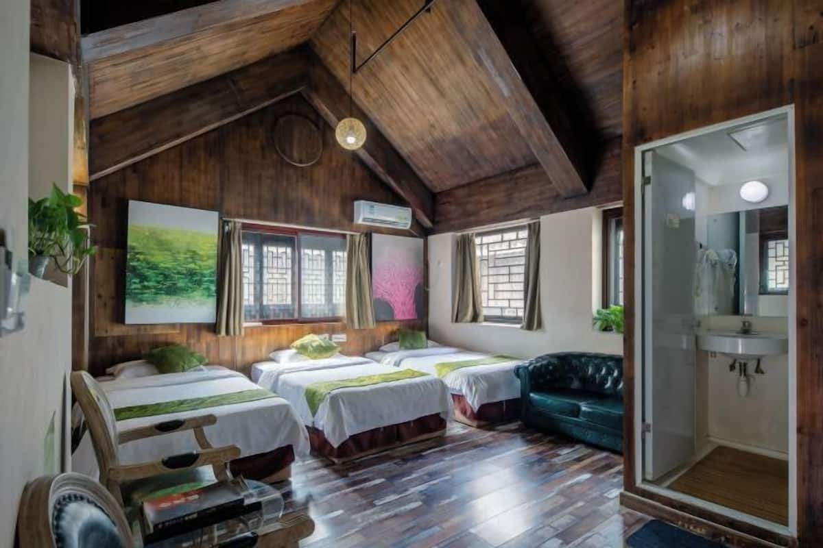 Quarto com vigas de madeira, duas camas com roupa de cama verde, um sofá verde e um banheiro com pia e espelho.
