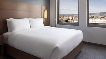 Quarto de hotel com uma cama grande, uma mesa de cabeceira, uma luminária na parede e uma janela com vista para a cidade.