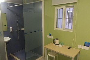 Casa de banho com cabine de duche em vidro, lavatório, janela e uma pequena mesa com artigos de higiene pessoal.