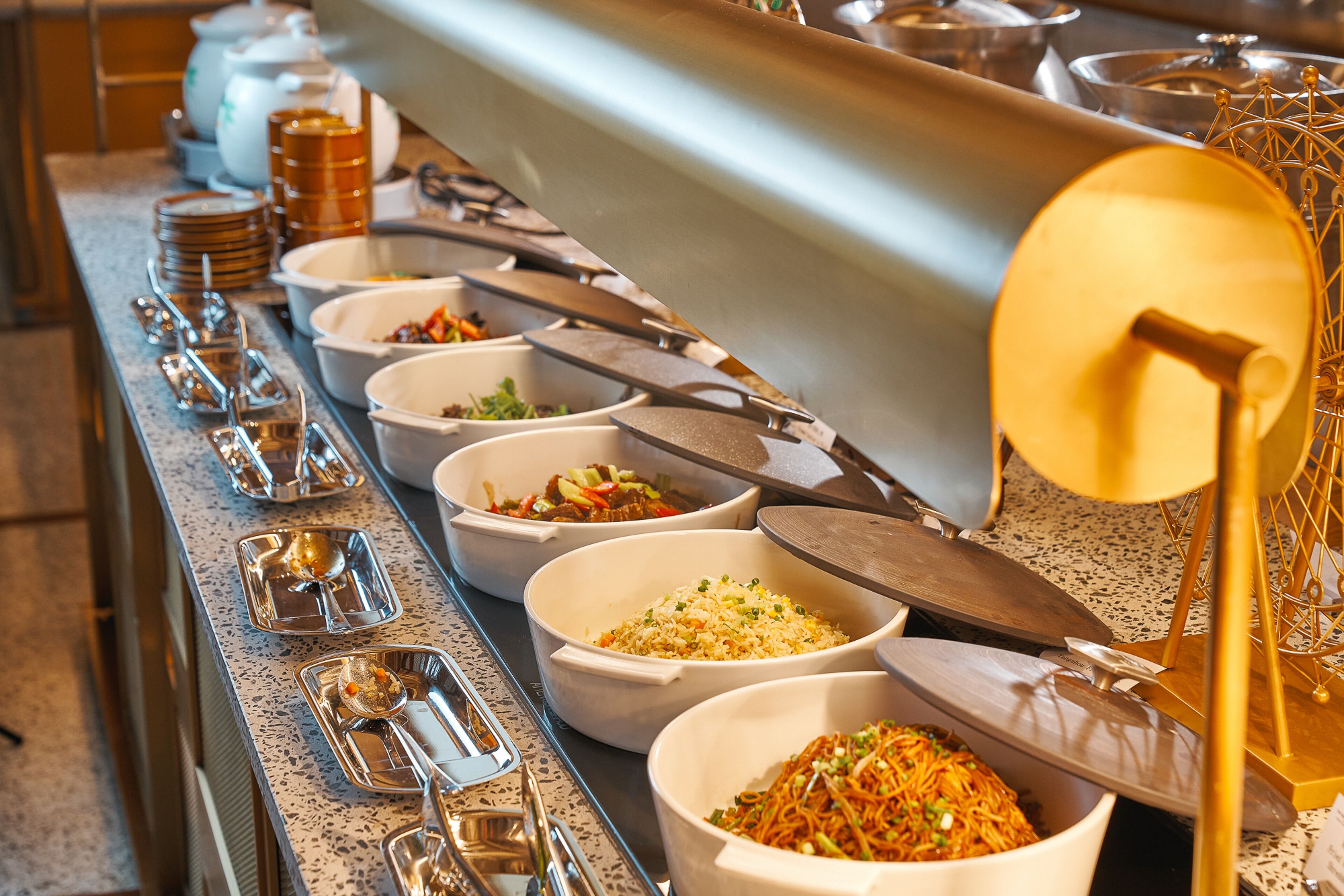 Uma mesa de buffet com diversos pratos, utensílios de servir e um carrinho dourado.