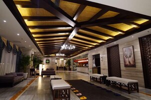 Um hall de hotel com teto alto, vigas de madeira e um lustre. Há áreas de estar com mesas e cadeiras.