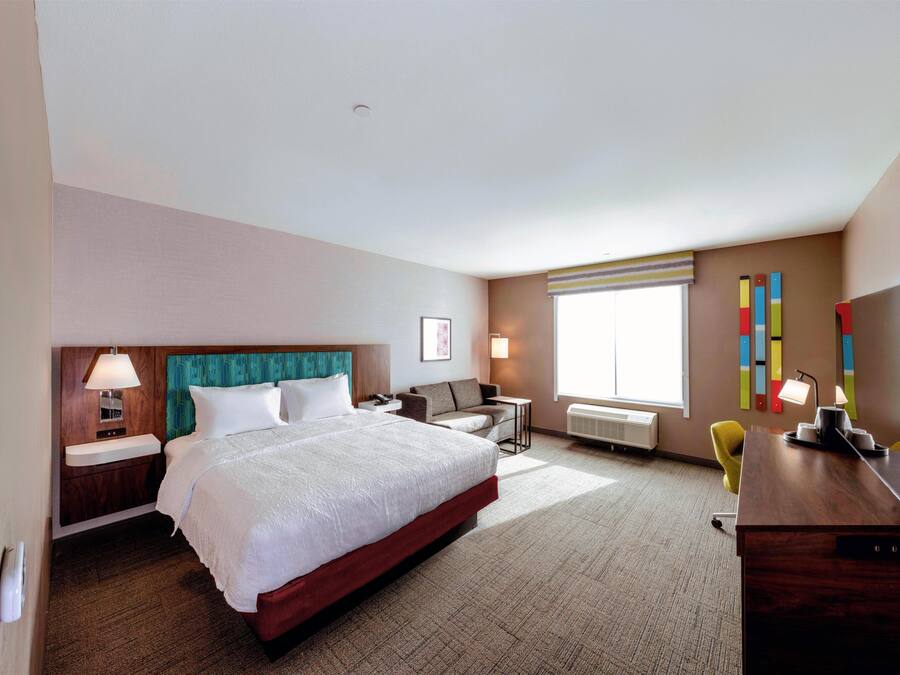 Quarto de hotel com uma cama grande, uma escrivaninha, uma cadeira, um sofá, uma televisão, um abajur e uma janela.