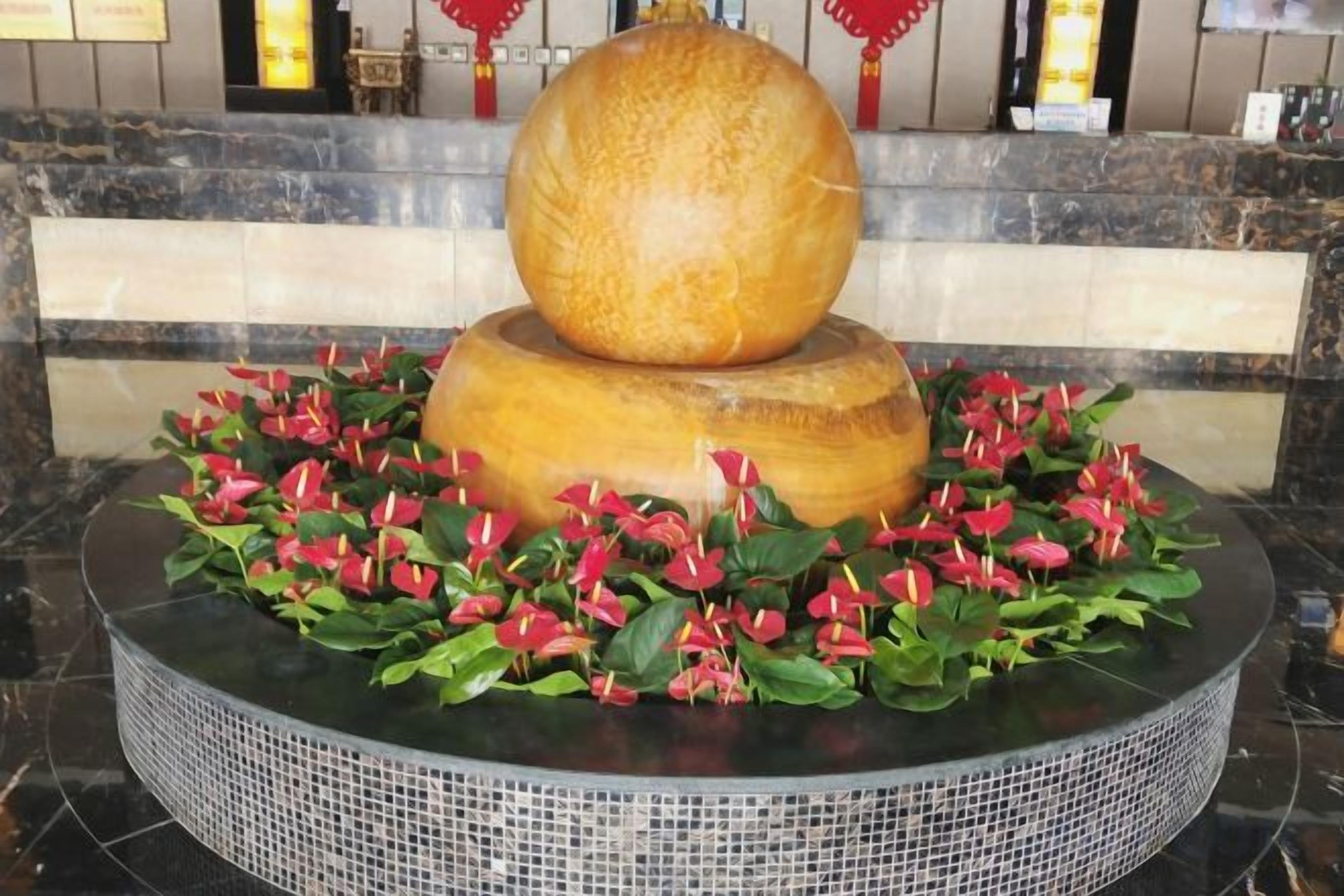 Uma fonte circular com uma grande esfera no topo, rodeada por flores vermelhas e vegetação.