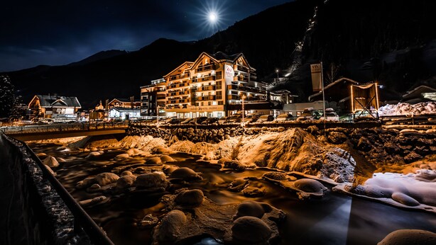 Um hotel com vários andares e uma placa chamativa, localizado às margens de um rio com pedras e bancos de neve.