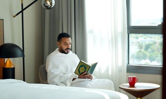 Uma pessoa lendo um livro em um quarto bem iluminado, com uma cama, uma mesinha com uma xícara e vista para o verde lá fora.