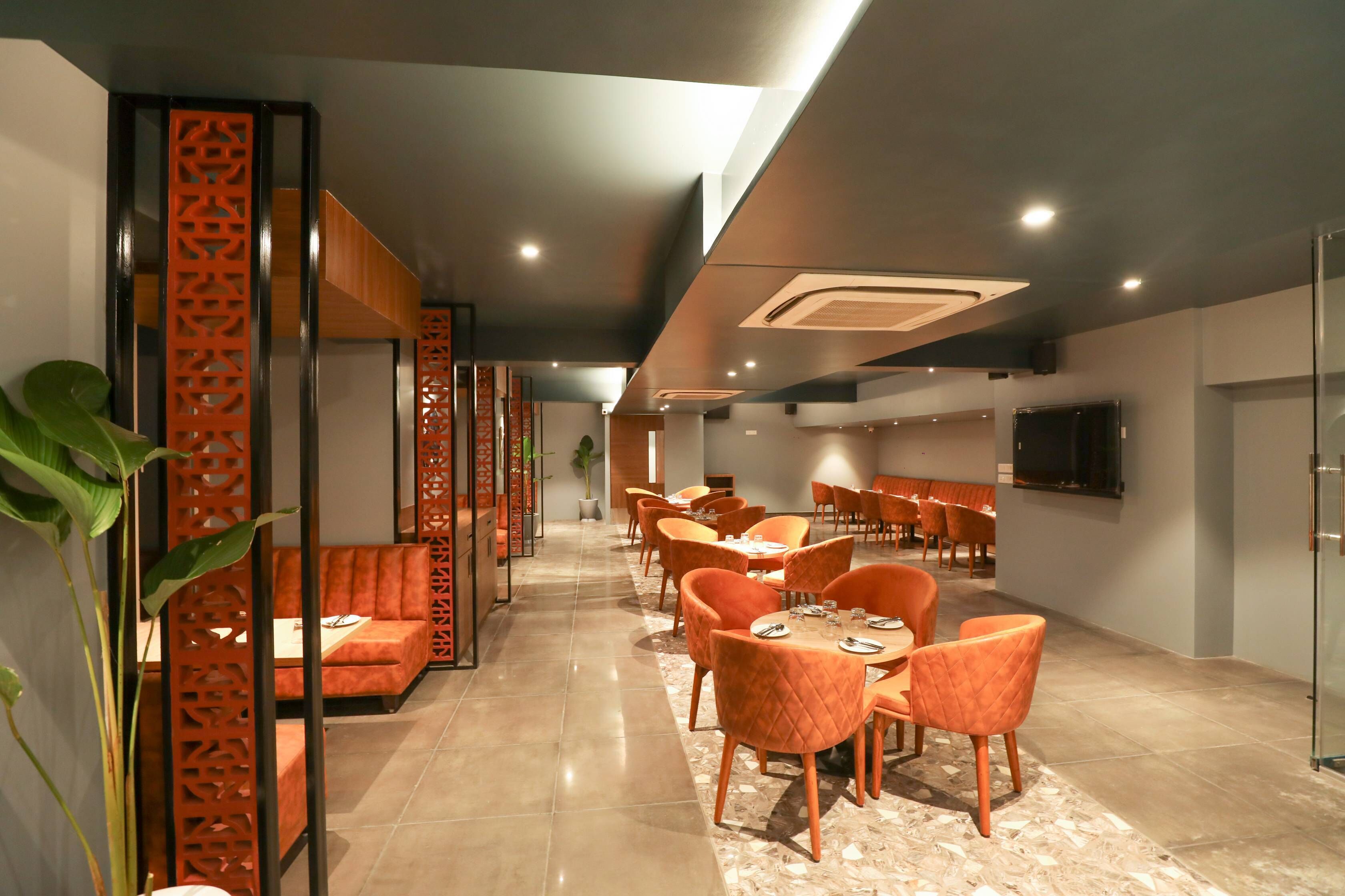 Um hall de hotel moderno com assentos laranja, uma área de refeições com mesas redondas e uma televisão na parede.