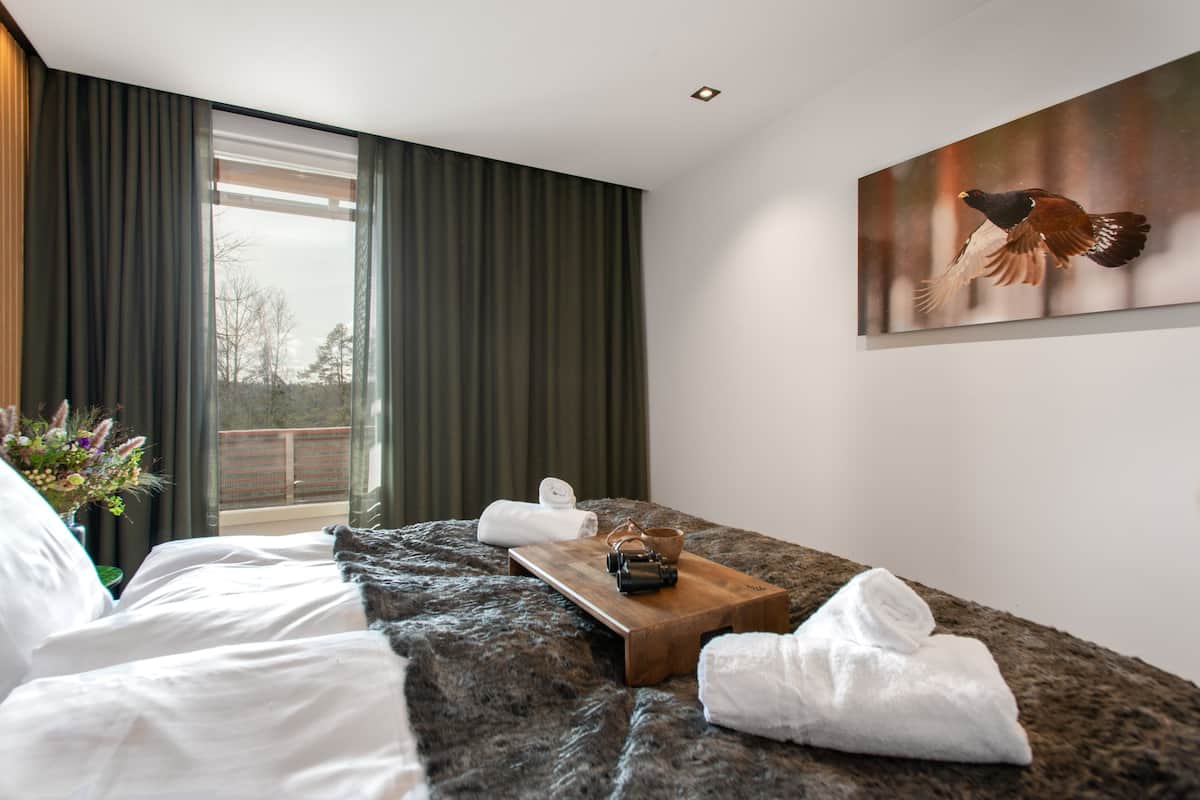 Quarto de hotel com uma cama grande, uma mesa de cabeceira de madeira, uma câmera e um quadro de um pássaro na parede.