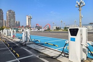 Uma área de estacionamento com estações de carregamento para veículos elétricos, céu claro e edifícios ao longe.
