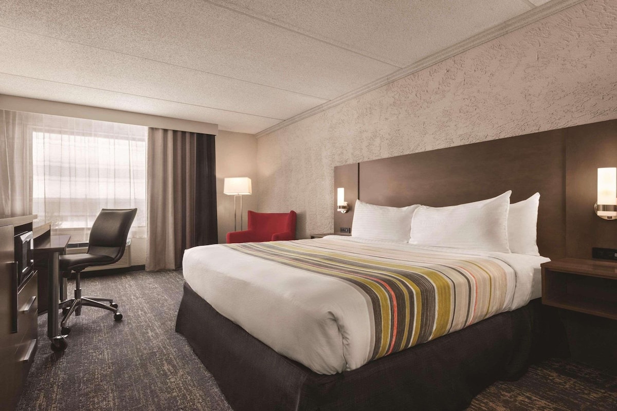 Quarto de hotel com uma cama grande, uma escrivaninha com cadeira, uma cadeira vermelha e um abajur.