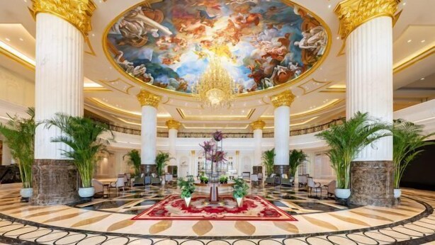 Um hall imponente com um grande mural, um candelabro e um arranjo floral central.