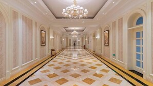 Um corredor longo e elegante, com chão quadriculado, lustres e obras de arte emolduradas nas paredes.