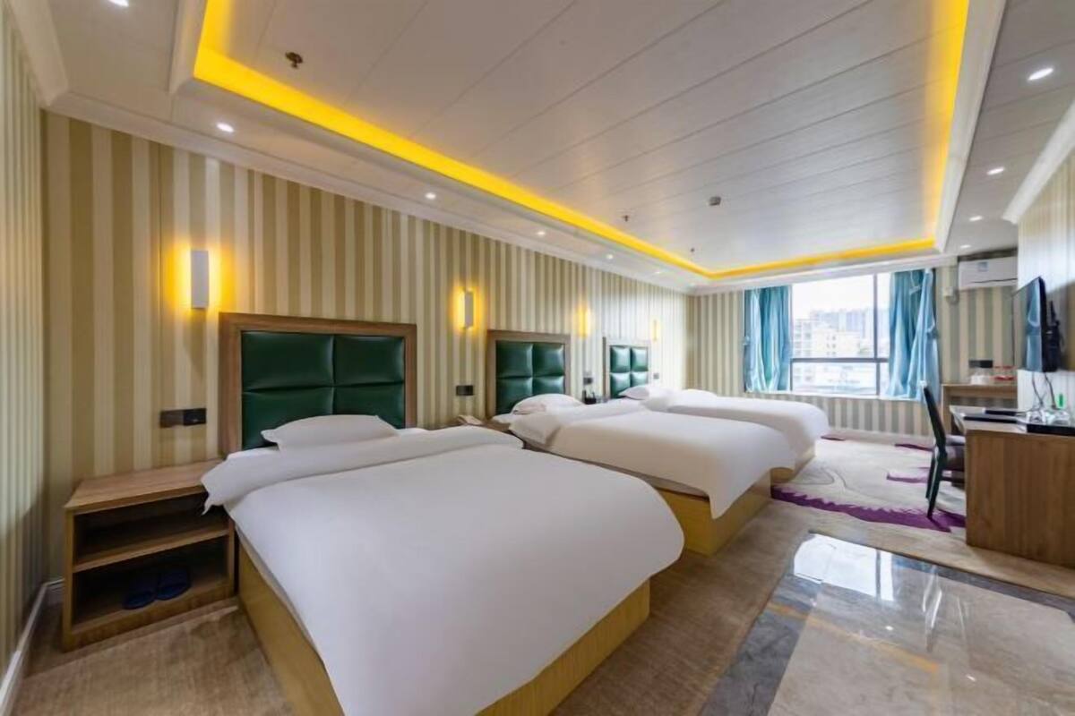 Quarto de hotel com três camas, uma escrivaninha, uma cadeira e janela com cortinas.