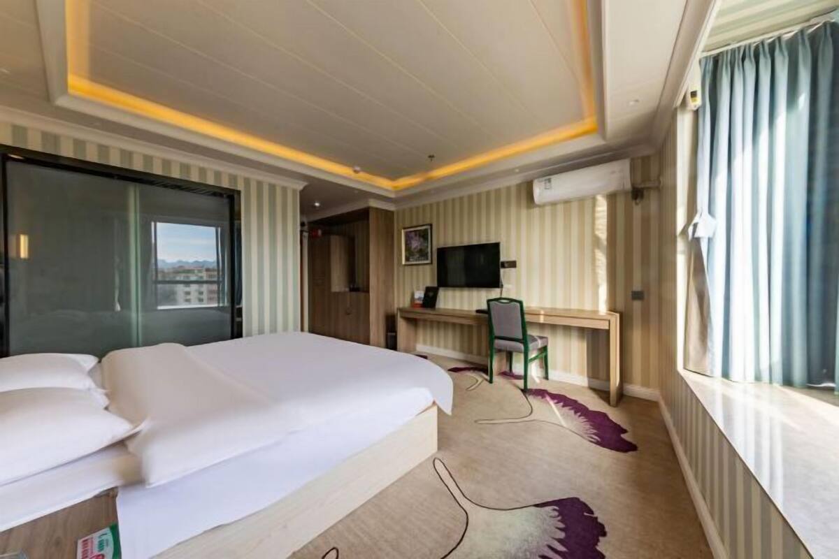 Quarto de hotel com cama, escrivaninha, cadeira, televisão e janela com cortinas.