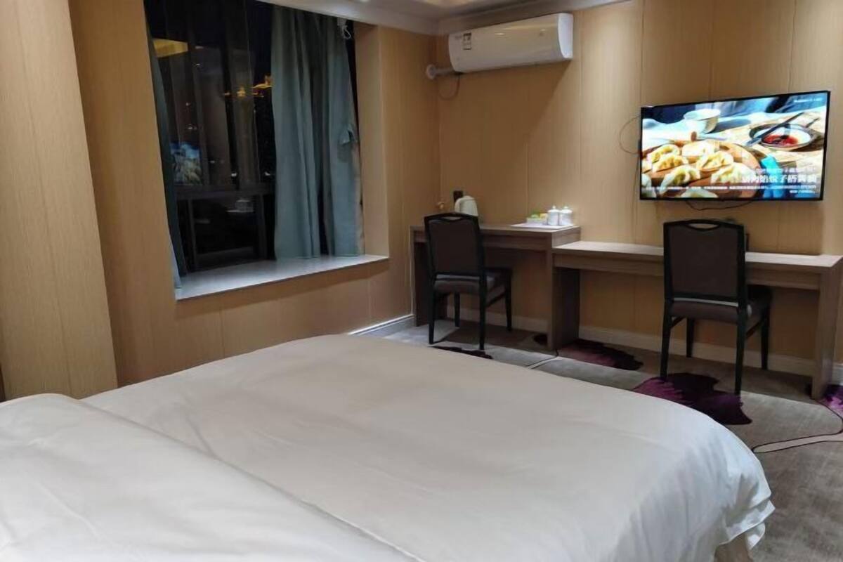 Quarto de hotel com cama, televisão, escrivaninha e duas cadeiras, além de janela com cortinas.
