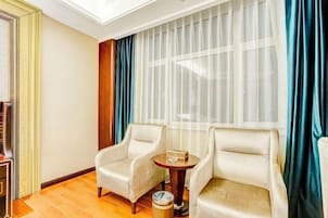 Quarto de hotel com duas poltronas bege, uma mesa redonda pequena e uma janela com cortinas brancas translúcidas e um tecido azul.