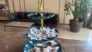Uma mesa de jantar com um arranjo decorativo central, incluindo um suporte escalonado com topo dourado e base azul, cercado por alimentos embalados.